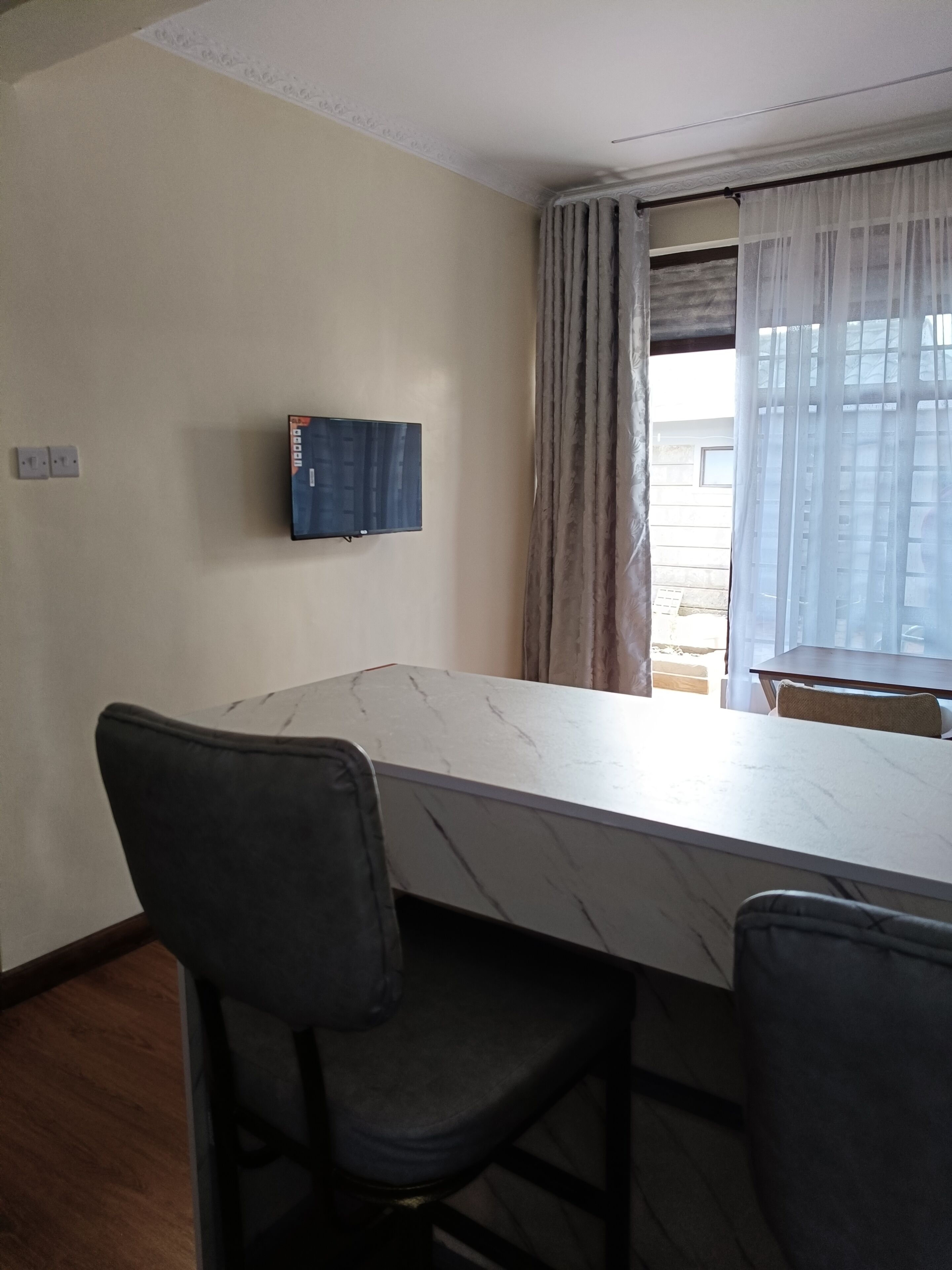 Um quarto com uma mesa de mármore, duas cadeiras, uma televisão na parede e uma janela com cortinas.