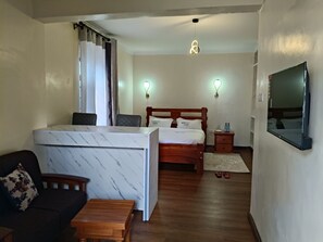 Quarto de hotel compacto com cama, sofá, mesa pequena, escrivaninha e televisão fixada na parede.