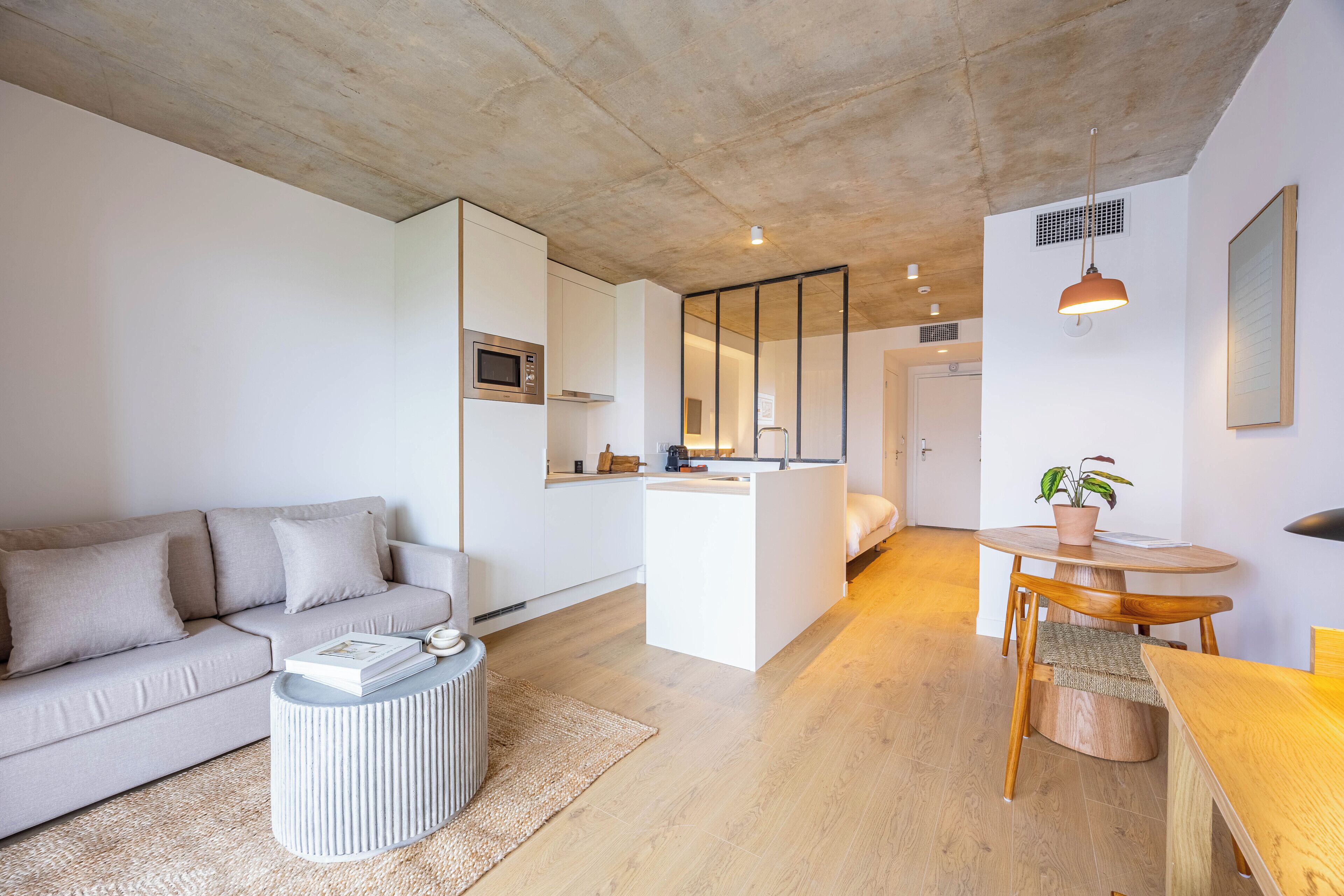 Um espaço de estar moderno com sofá, mesa de centro redonda, área de jantar com mesa e cadeiras de madeira, cozinha com micro-ondas e um espelho grande.
