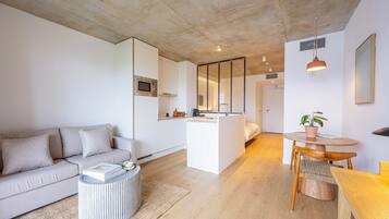 Um espaço de estar moderno com sofá, mesa de centro redonda, área de jantar com mesa e cadeiras de madeira, cozinha com micro-ondas e um espelho grande.