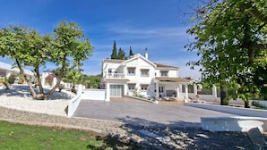 Uma casa de dois andares com entrada por automóvel, fachada branca e vegetação ao redor.