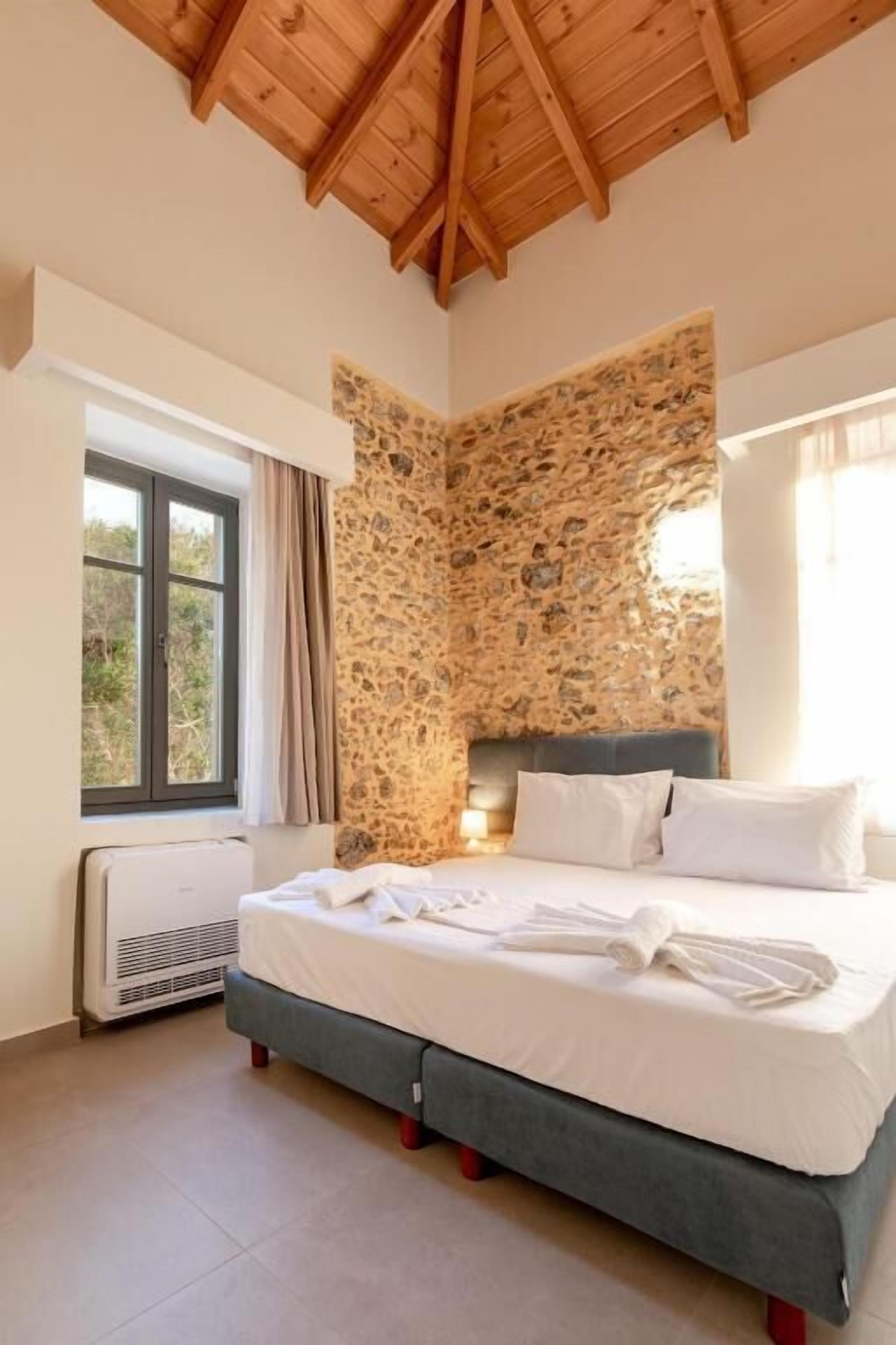 Um quarto com parede de pedra, cama com roupa de cama branca, teto de madeira e janela com cortinas.