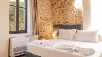 Um quarto com parede de pedra, cama com roupa de cama branca, teto de madeira e janela com cortinas.