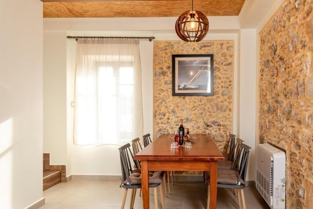 Uma área de jantar com mesa de madeira, cadeiras, um quadro pendurado na parede, uma janela com cortinas e uma parede de pedra.