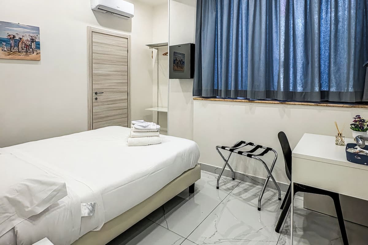 Quarto de hotel com cama, escrivaninha, cadeira, mesa pequena com uma garrafa e uma caixa, espelho, janela com cortinas e ar condicionado.