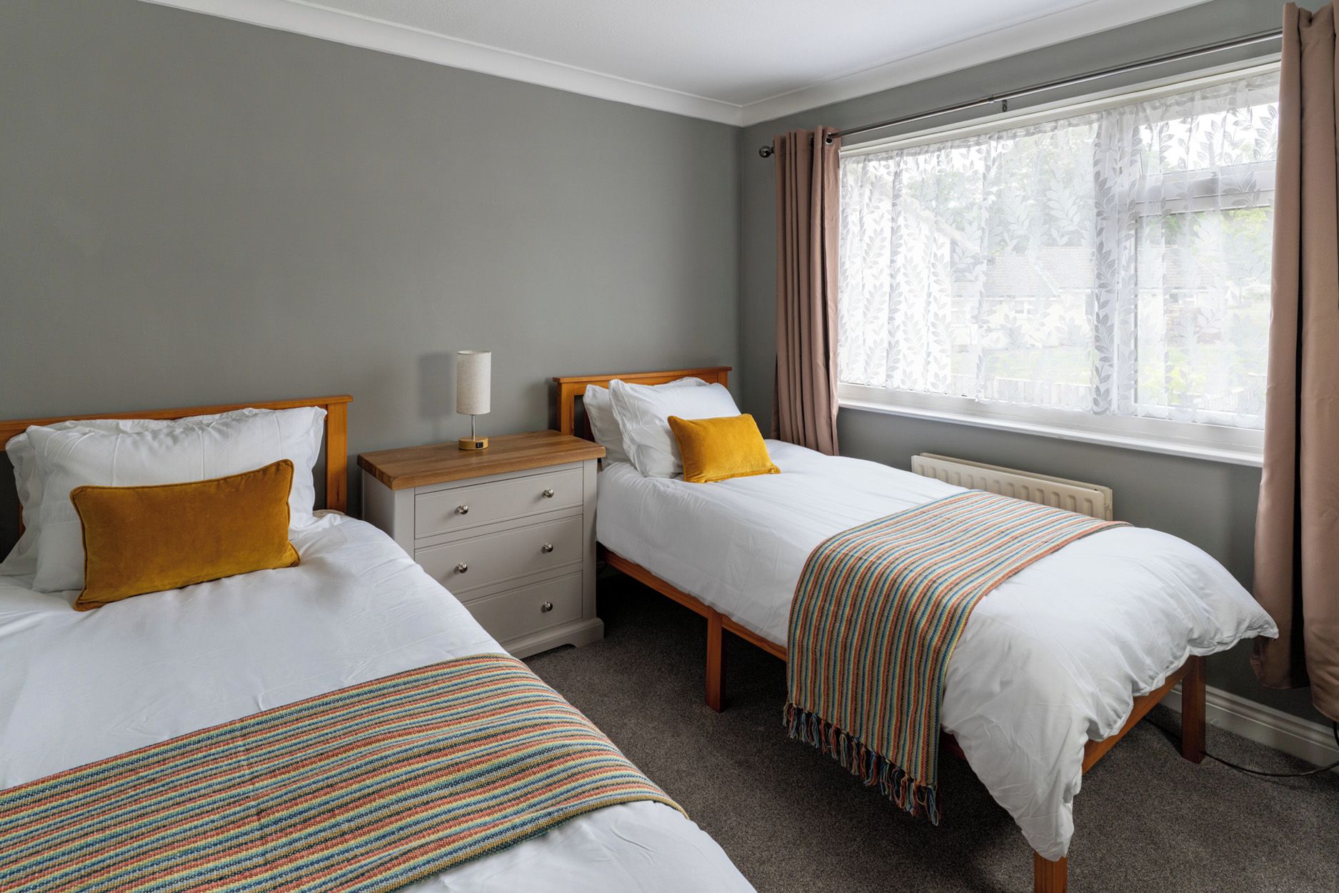 Quarto com duas camas, mesa de cabeceira, luminária e janela com cortinas.