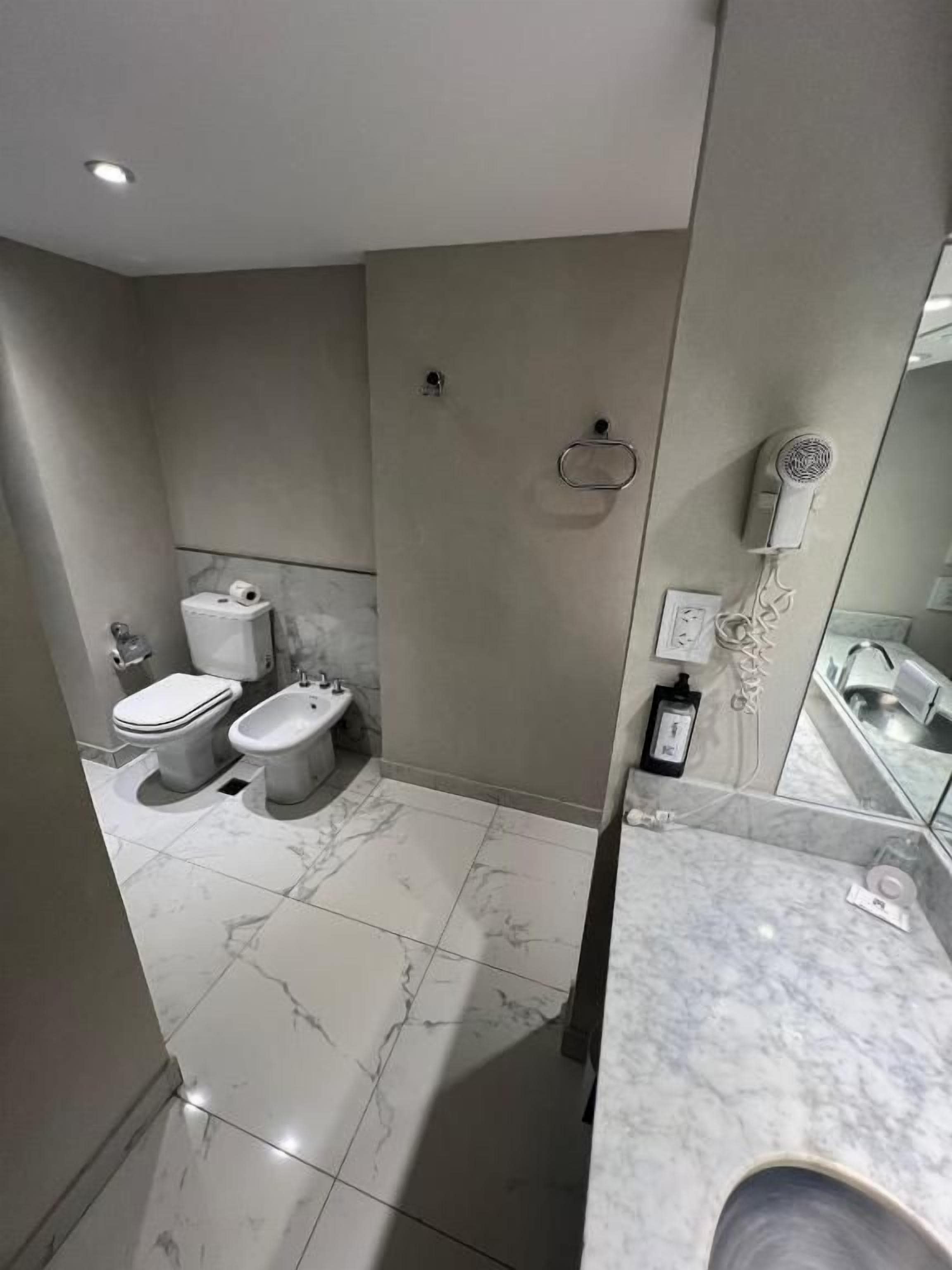 Casa de banho com sanita, bidé e secador de cabelo fixado na parede.