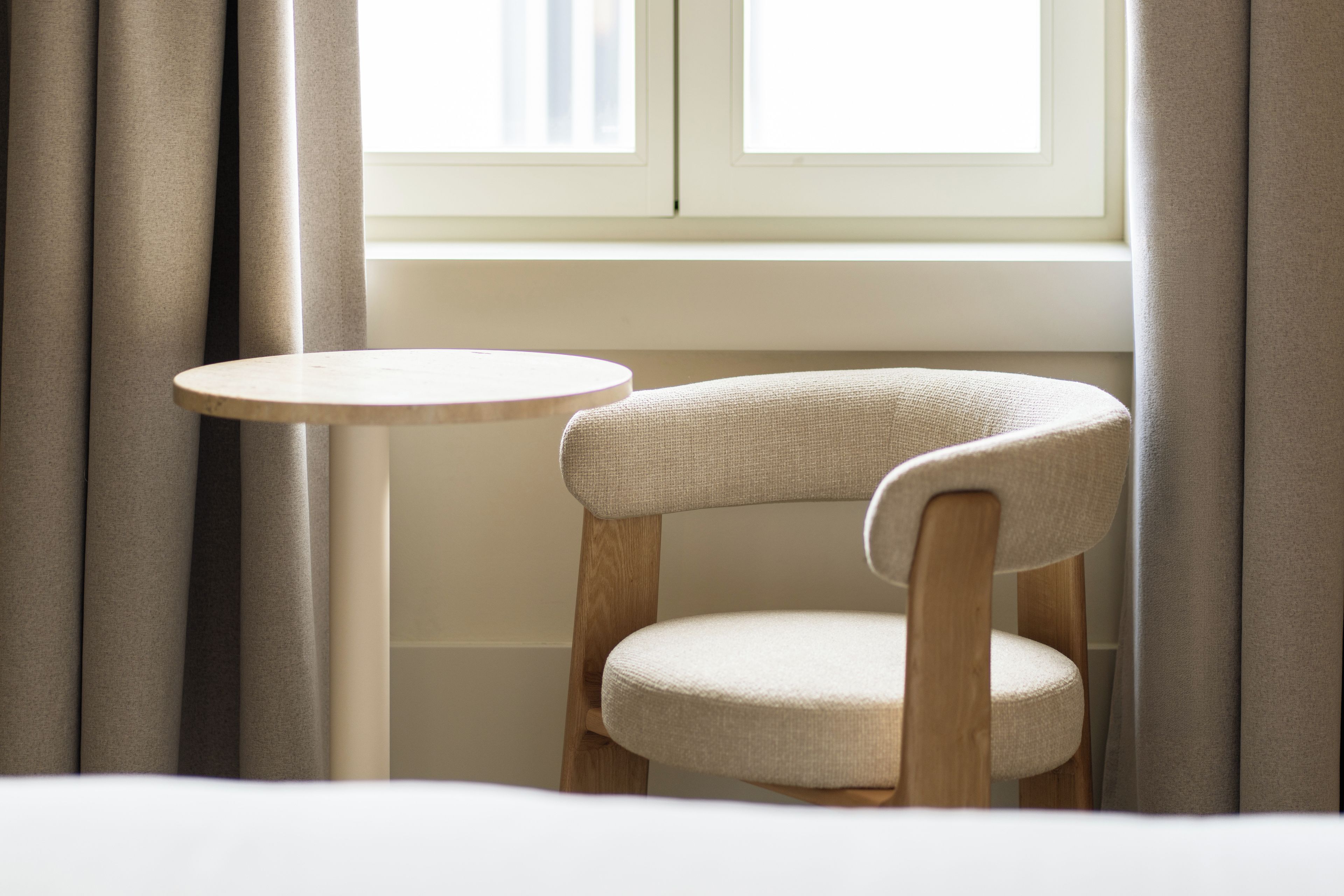 Uma cadeira com estrutura de madeira e assento acolchoado, colocada ao lado de uma pequena mesa redonda, com cortinas entreabertas mostrando uma janela com persianas.
