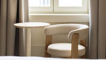 Uma cadeira com estrutura de madeira e assento acolchoado, colocada ao lado de uma pequena mesa redonda, com cortinas entreabertas mostrando uma janela com persianas.