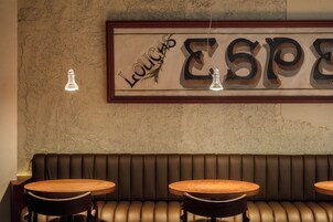 Interior de restaurante com área de estar em bancos de madeira, duas mesas redondas e uma placa na parede com os dizeres 'Lounges' e 'Espresso'.