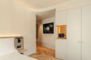 Quarto de hotel moderno com cama, uma mesinha, guarda-roupa e televisão fixada na parede.