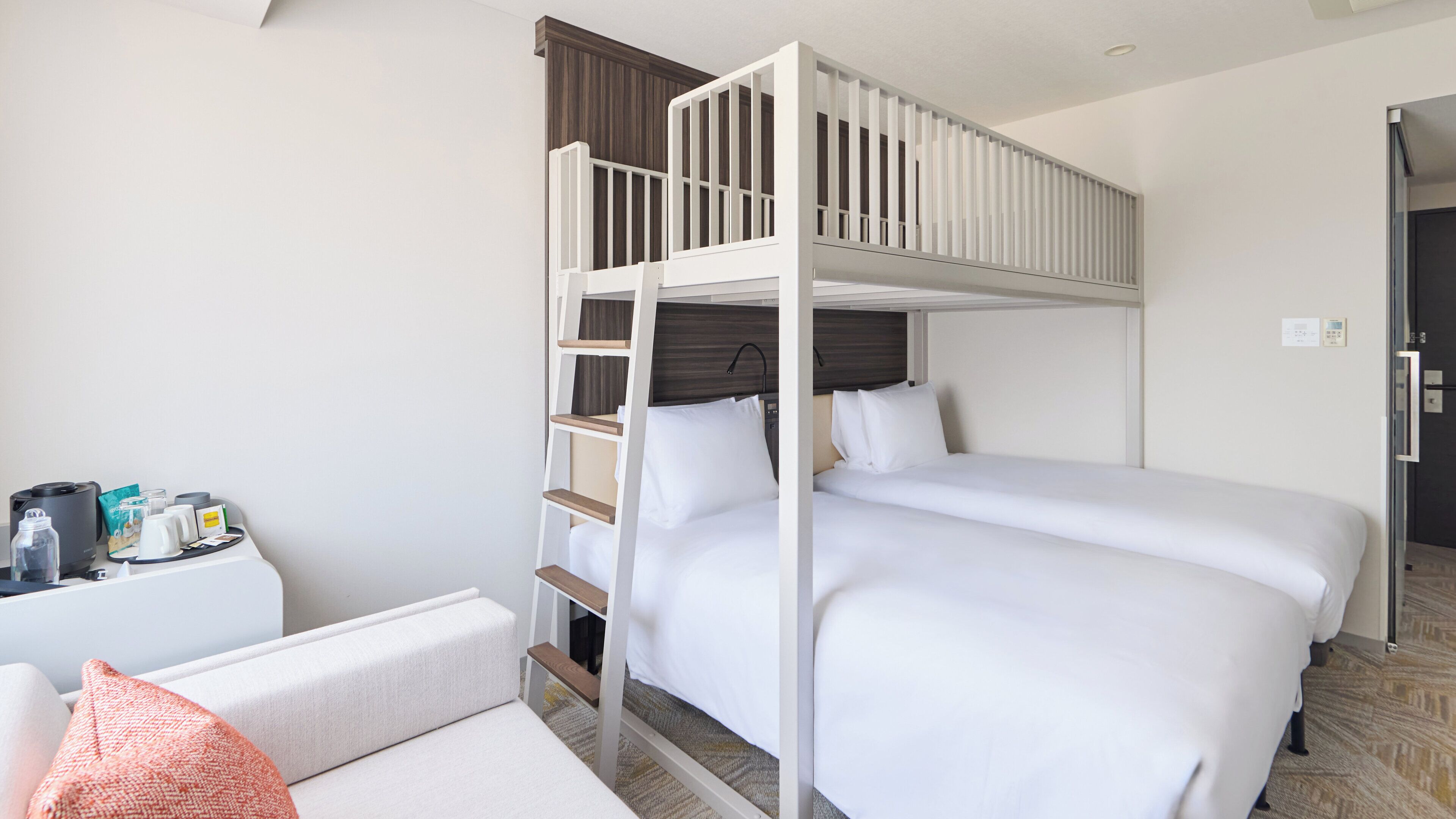 Quarto de hotel com beliche, uma mesa com cafeteira e uma cadeira.
