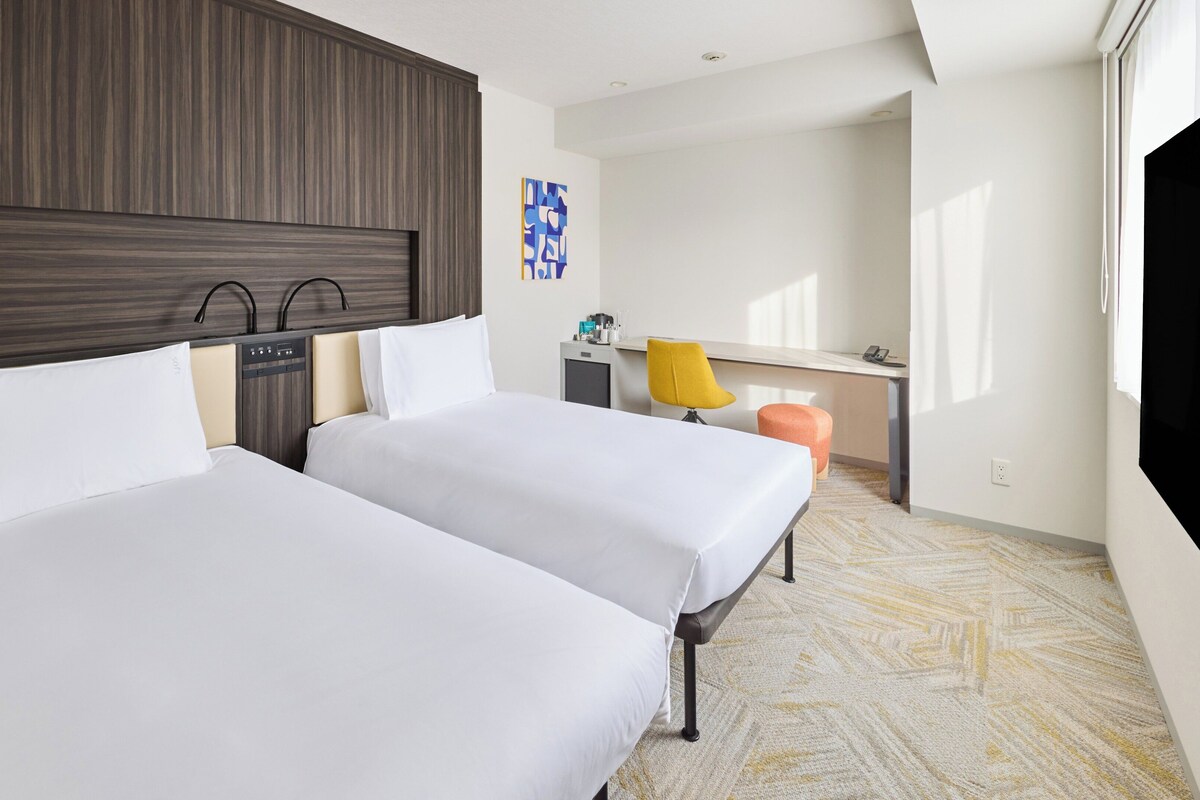 Quarto de hotel com duas camas, uma escrivaninha, uma cadeira e uma televisão.