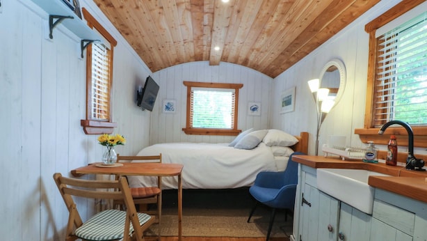 Quarto compacto com cama, uma mesinha com cadeira, pia e janela com persianas.