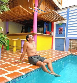 Um homem sentado à beira da piscina, sob uma pérgola de madeira, com área de bar e um sinal indicando saída de emergência.