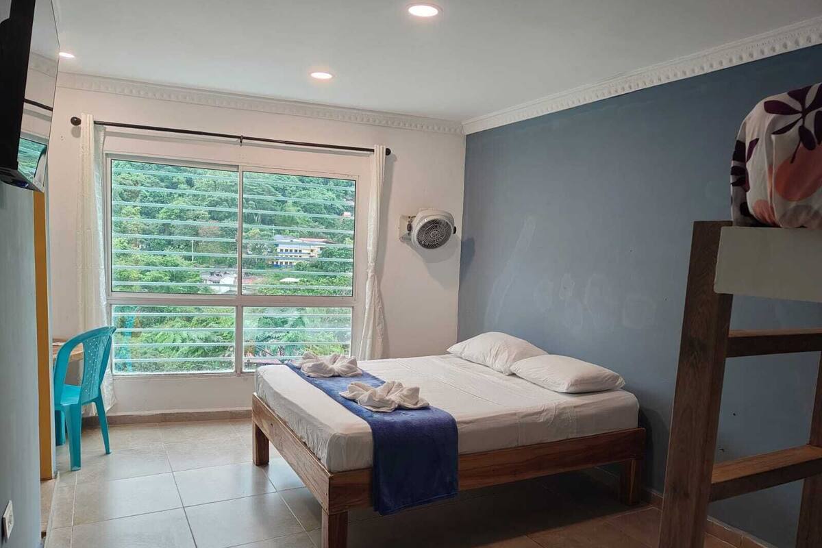 Quarto com cama, janela com persianas, ventilador e uma cadeira azul.