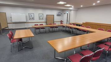 Uma sala de conferências com mesas compridas e cadeiras vermelhas, quadro branco e quadros nas paredes.