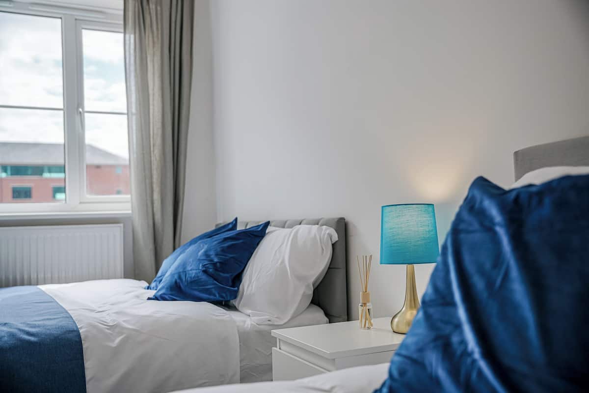 Quarto de hotel com cama, mesa de cabeceira, luminária e janela com cortinas.