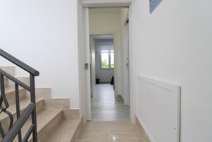 Um corredor com escadas que levam a outro cômodo, paredes brancas e uma porta no final.