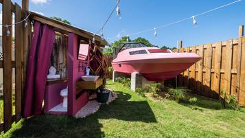 Um barco rosa atracado em um quintal com cerca de madeira, um pequeno galpão e uma guirlanda de luzes.