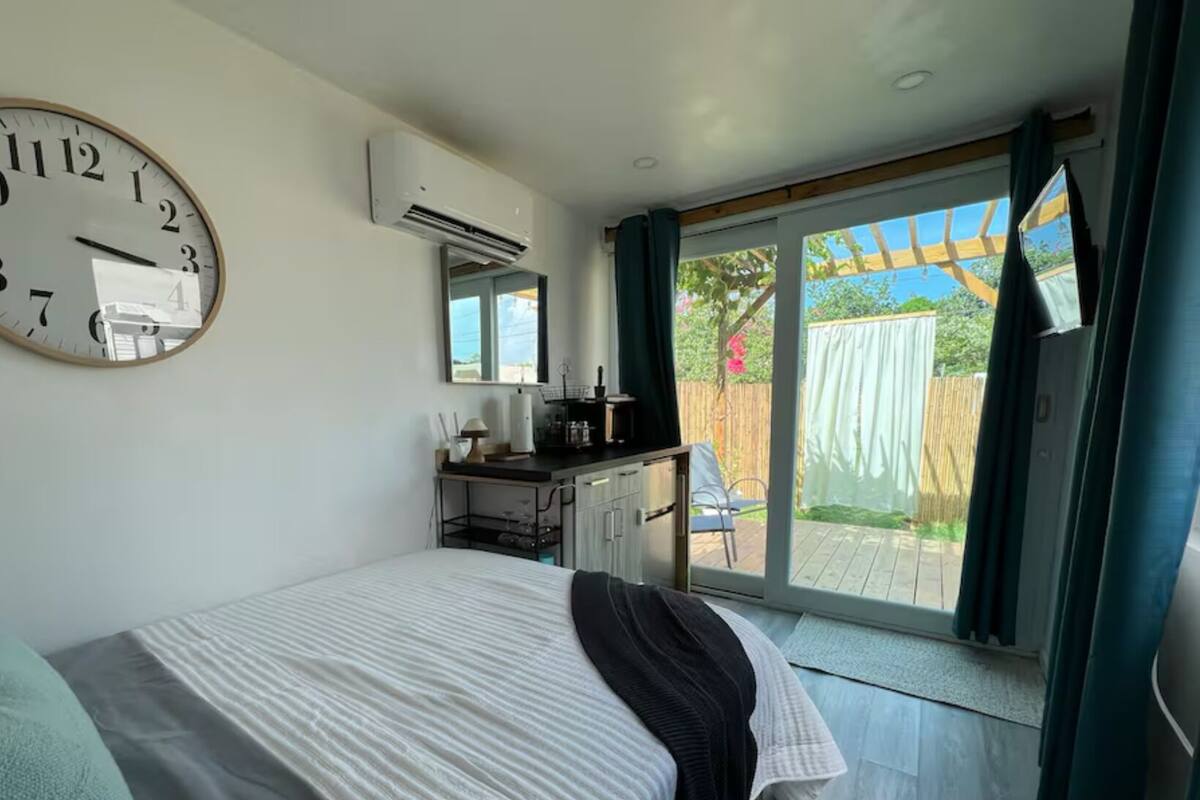 Quarto com cama, relógio, escrivaninha e porta de vidro deslizante para uma varanda.