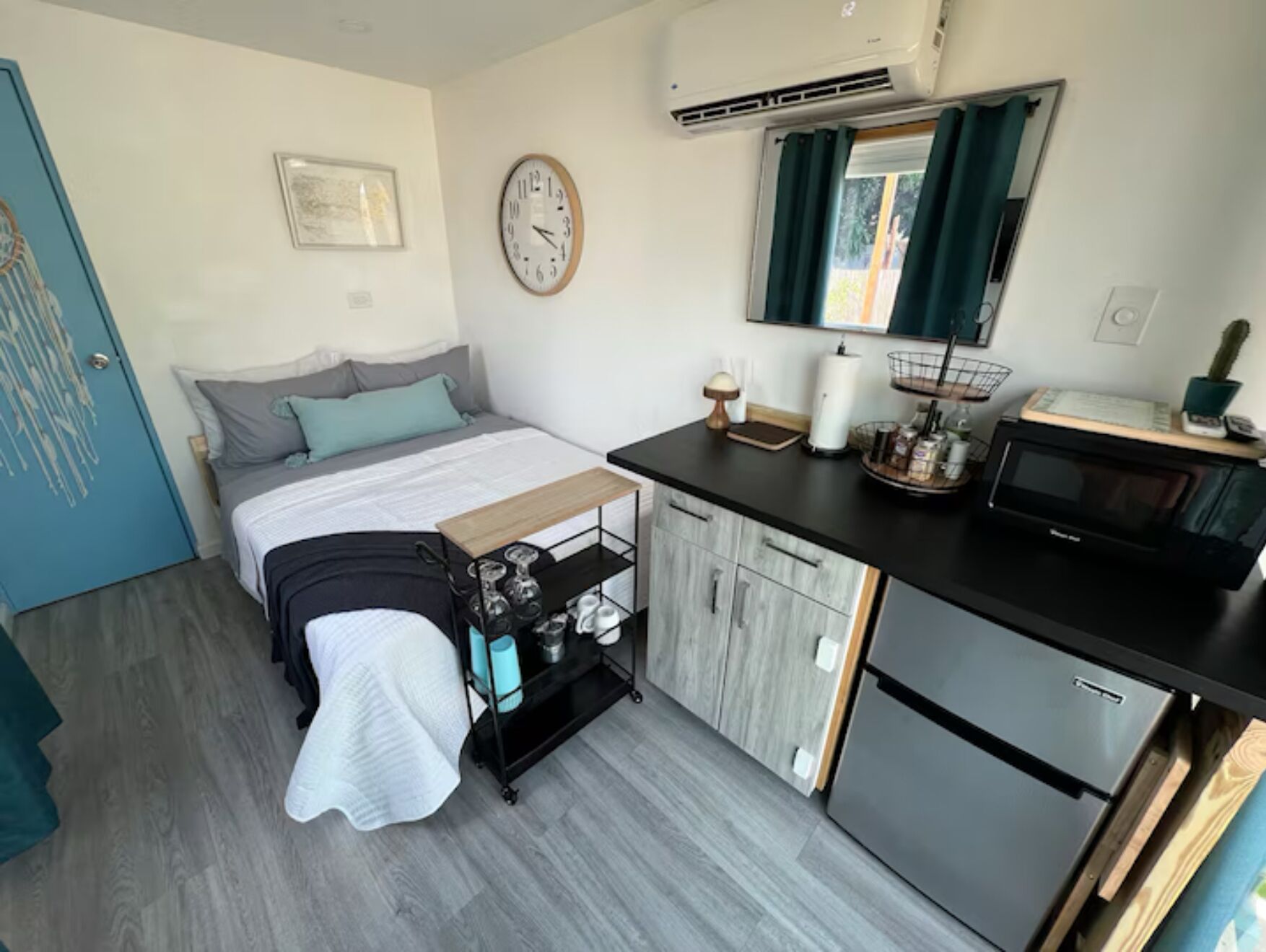 Quarto compacto com cama, pequeno espaço de cozinha, relógio e janela com cortinas.