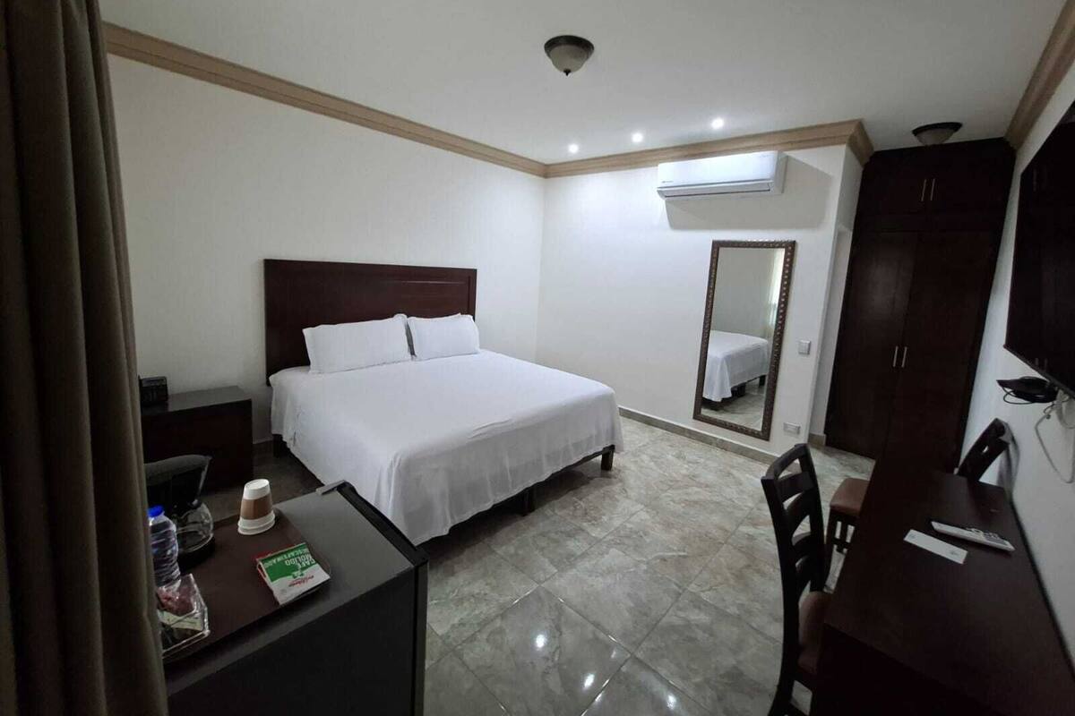 Quarto de hotel com cama, escrivaninha, cadeira, espelho, guarda-roupa e ar condicionado.