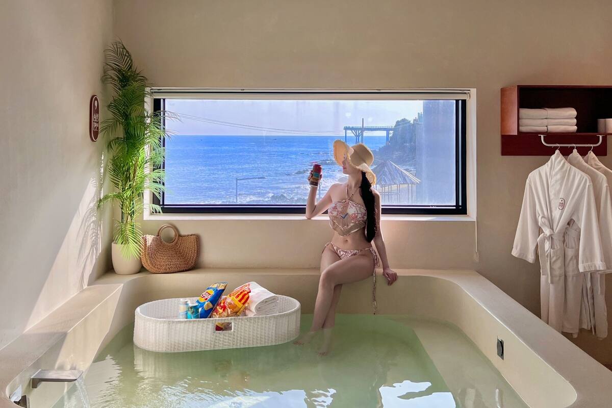 Quarto com uma janela grande, banheira, planta em vaso e prateleira com toalhas.