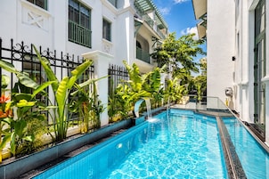 Piscina ao ar livre com superfície de água azul cristalina, cercada por vegetação tropical exuberante e um edifício branco com molduras de janelas verdes.