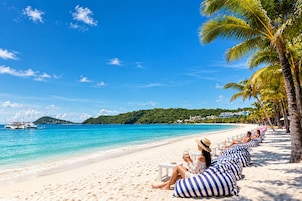 Uma praia com areia branca, águas azul-claras e palmeiras. Há cadeiras de praia e guarda-sóis ao longo da costa.