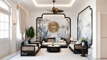 Uma sala de estar ampla com teto alto, ventilador de teto e um grande mural na parede. Há dois sofás, uma mesa de centro e uma mesa lateral com um vaso de flores.