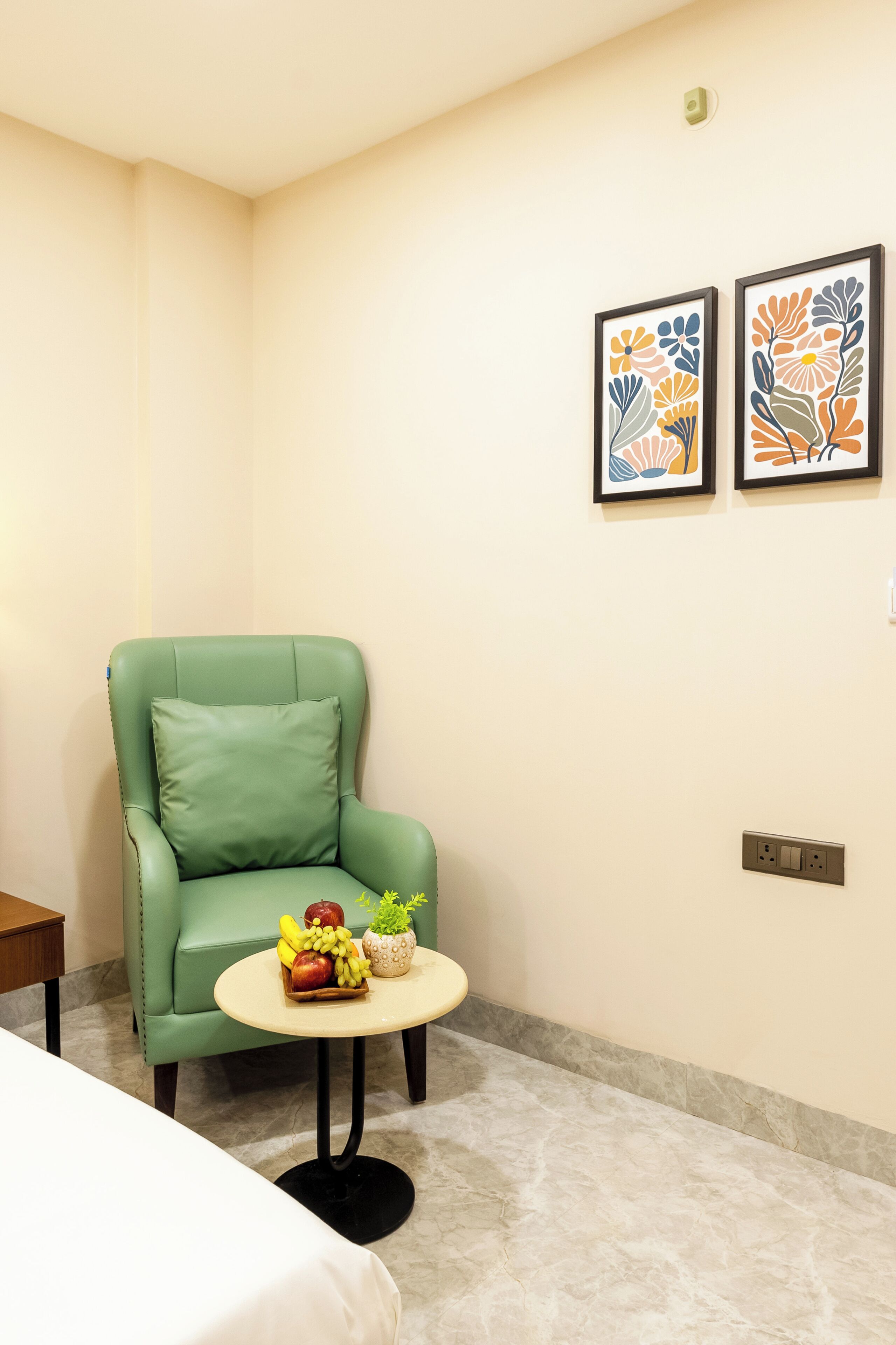 Um quarto com uma poltrona verde, uma mesinha redonda com frutas e uma planta, e duas obras de arte com flores emolduradas na parede.