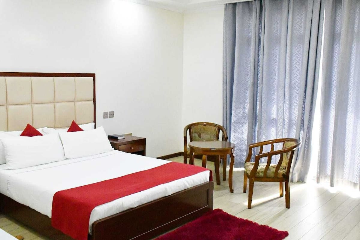 Quarto de hotel com uma cama, duas cadeiras, uma mesinha e uma janela com cortinas.