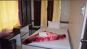 Uma cama de solteiro com colchão branco, manta floral vermelha e travesseiro branco. Há uma pequena mesa de cabeceira com abajur, lixeira e janela com cortinas.