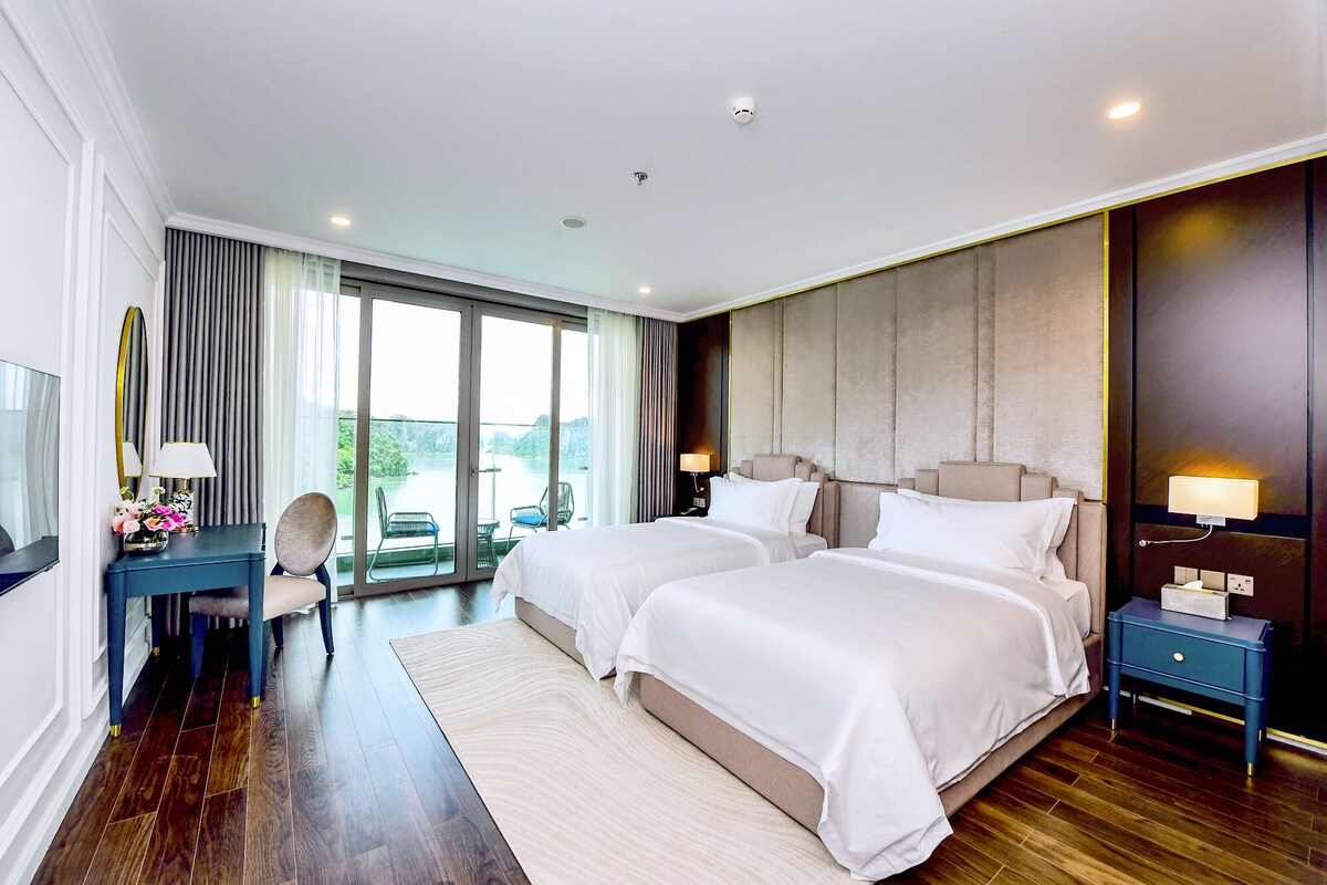 Quarto de hotel com duas camas, uma mesa com cadeira, televisão e uma janela grande com vista.