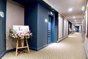 Um corredor de hotel com um arranjo floral sobre um cavalete, uma porta com uma placa e obras de arte emolduradas em cavaletes.