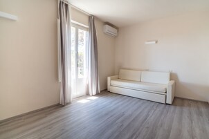 Um quarto com piso de madeira clara, um sofá branco, uma porta de vidro deslizante com cortinas e um aparelho de ar condicionado fixado na parede.