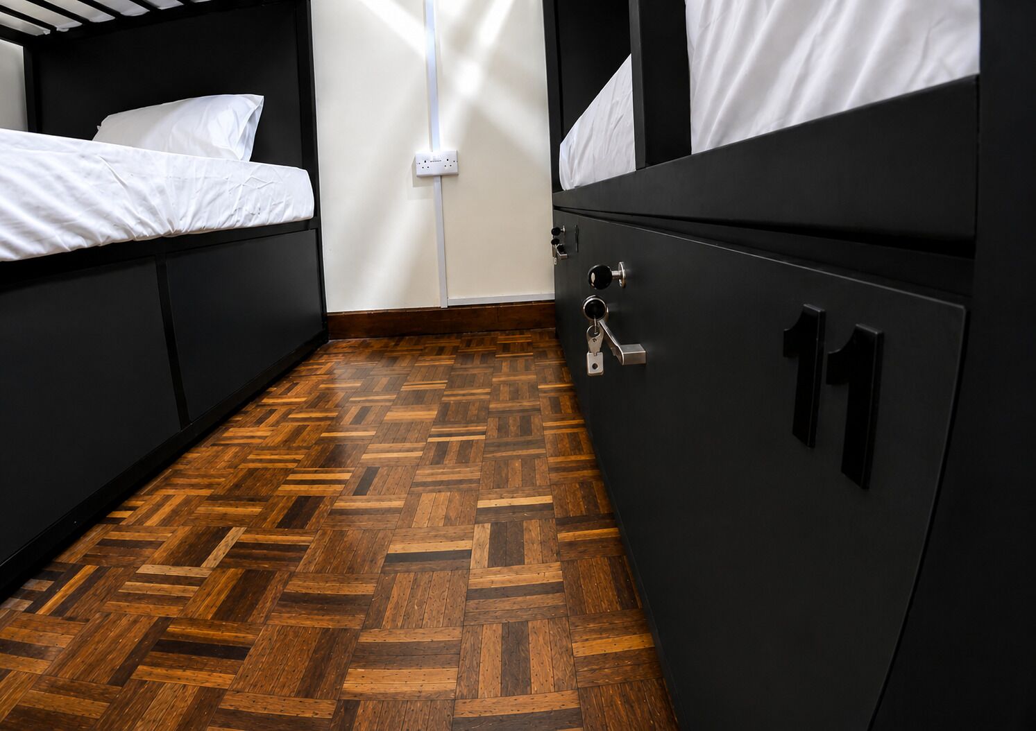 Quarto com duas camas, piso de parquet de madeira, parede branca com tomada e armário preto com fechadura.
