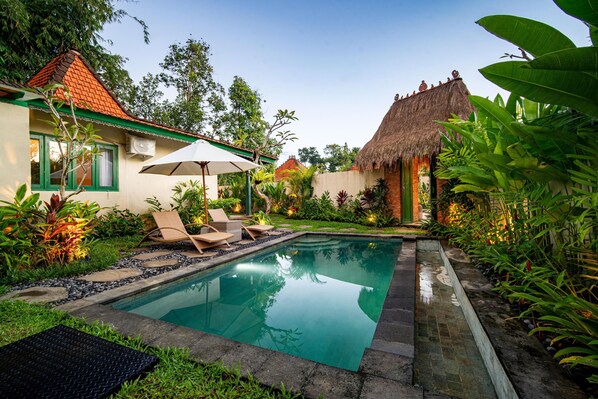 Uma piscina com espreguiçadeiras e um guarda-sol, rodeada por vegetação exuberante e arquitetura tradicional.
