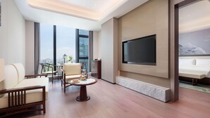 Quarto de hotel moderno com uma grande TV de tela plana fixada em uma parede texturizada, um sofá confortável, uma mesa de jantar com cadeiras e vista para a cidade através de amplas janelas.