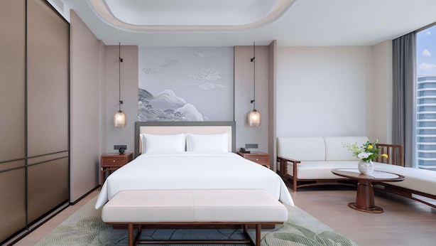 Quarto de hotel moderno com uma cama grande, um espaço de estar com uma mesa e vista para a cidade.