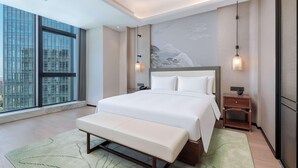 Quarto de hotel moderno com uma cama grande, um banco, uma mesa de cabeceira e vista da cidade.