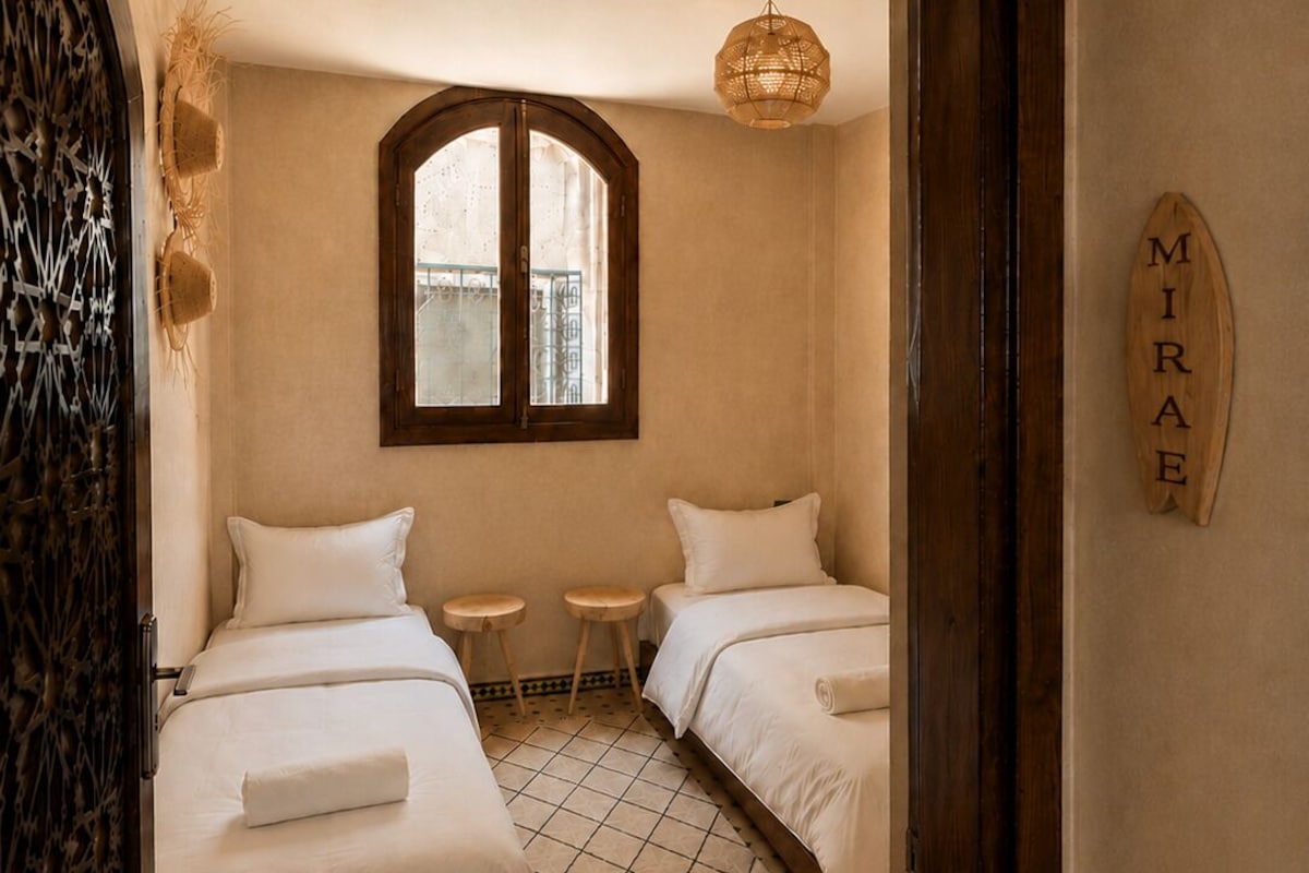 Quarto de hotel com duas camas, uma janela, uma porta de madeira e piso de azulejos.