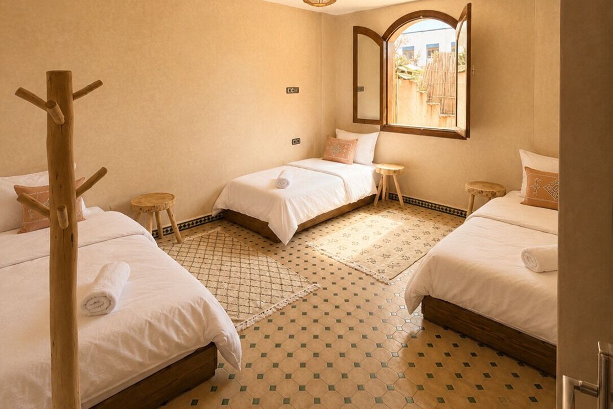 Quarto de hotel com duas camas, um cabideiro de madeira, um pequeno banquinho, piso estampado e um espelho grande.
