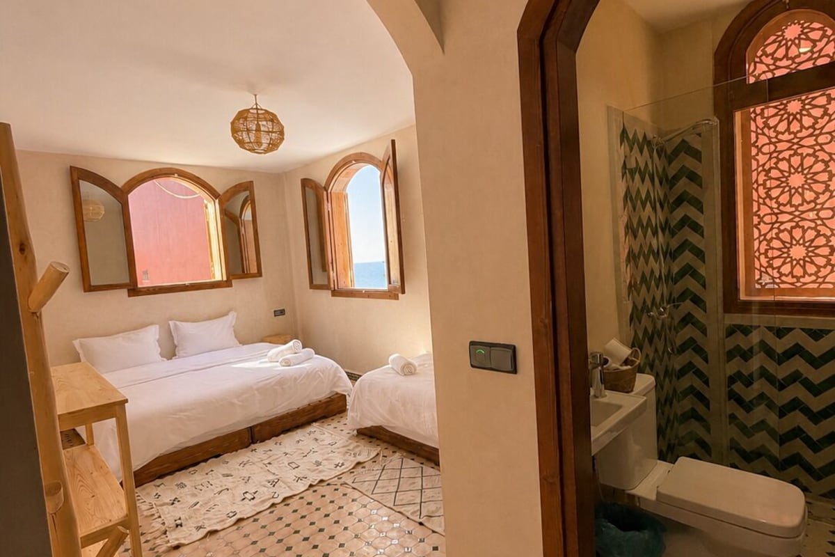 Quarto de hotel com uma cama, uma cadeira de madeira, uma mesa pequena, um espelho e um banheiro com vaso sanitário e chuveiro.