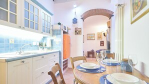 Uma área de refeições com mesa de madeira posta para quatro pessoas, cozinha com armários brancos e azulejos azuis, e um arco de tijolos que leva a outro cômodo.