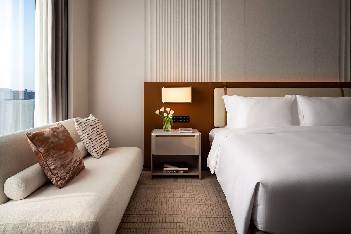 Quarto de hotel com cama, sofá, mesa de cabeceira com abajur e vista da cidade.