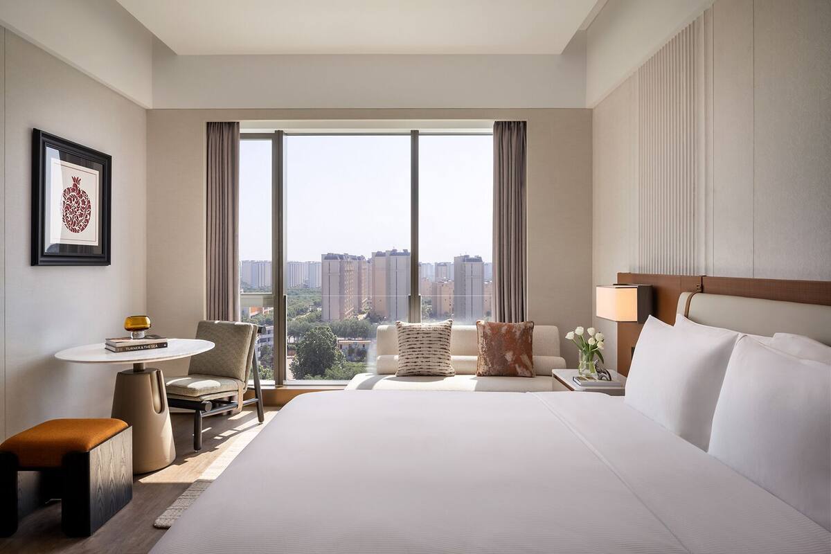 Quarto de hotel com uma cama grande, uma escrivaninha, uma cadeira, uma mesinha e vista para a paisagem urbana.