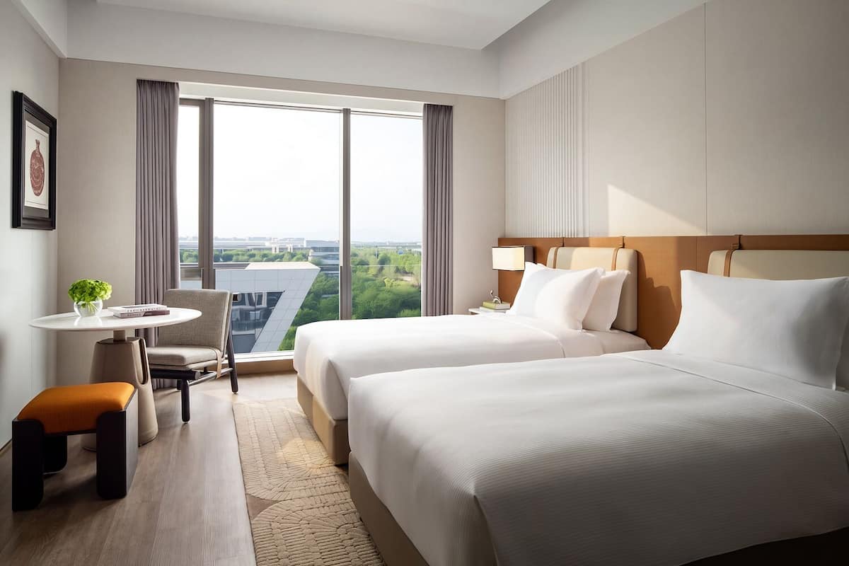 Quarto de hotel com duas camas, uma mesinha com uma planta, uma cadeira e vista para o verde através de amplas janelas.