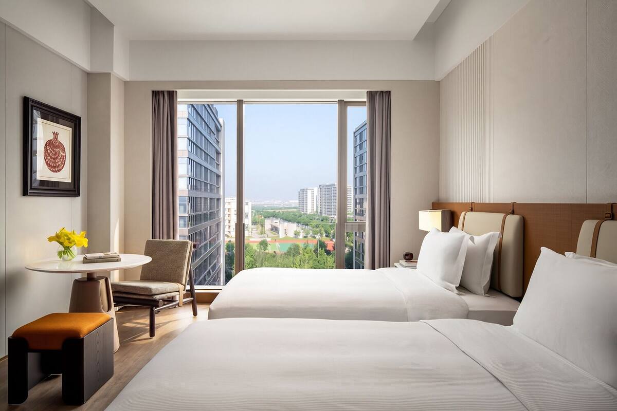 Quarto de hotel com duas camas, uma mesa de escritório, uma cadeira, uma pequena mesa com flores e grandes janelas com vista para a cidade.